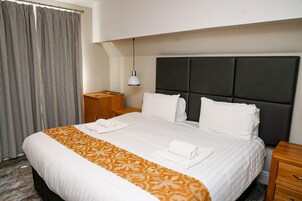 Quarto de hotel com uma cama grande, uma mesinha de cabeceira de madeira, uma escrivaninha e uma luminária.
