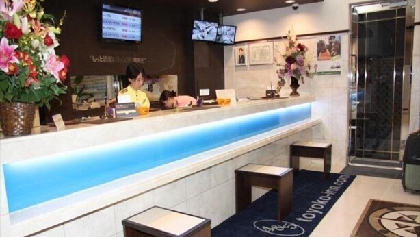 Uma área de recepção de hotel com um balcão azul, uma tela digital e um tapete com o site do hotel.