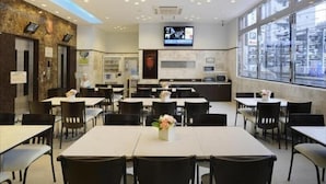 Um restaurante com várias mesas e cadeiras, uma tela de TV na parede e vista da cidade através de amplas janelas.