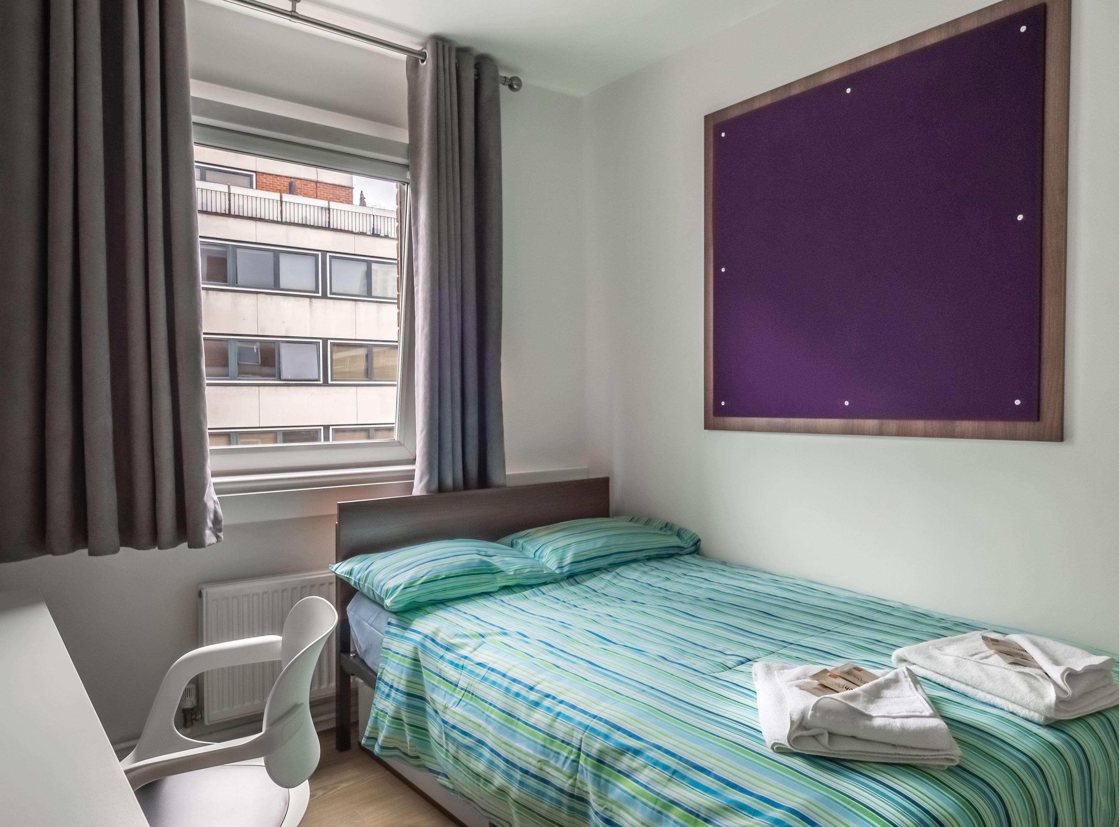 Quarto pequeno e bem arrumado, com uma cama de solteiro, uma cadeira, uma janela com cortinas e um mural na parede.