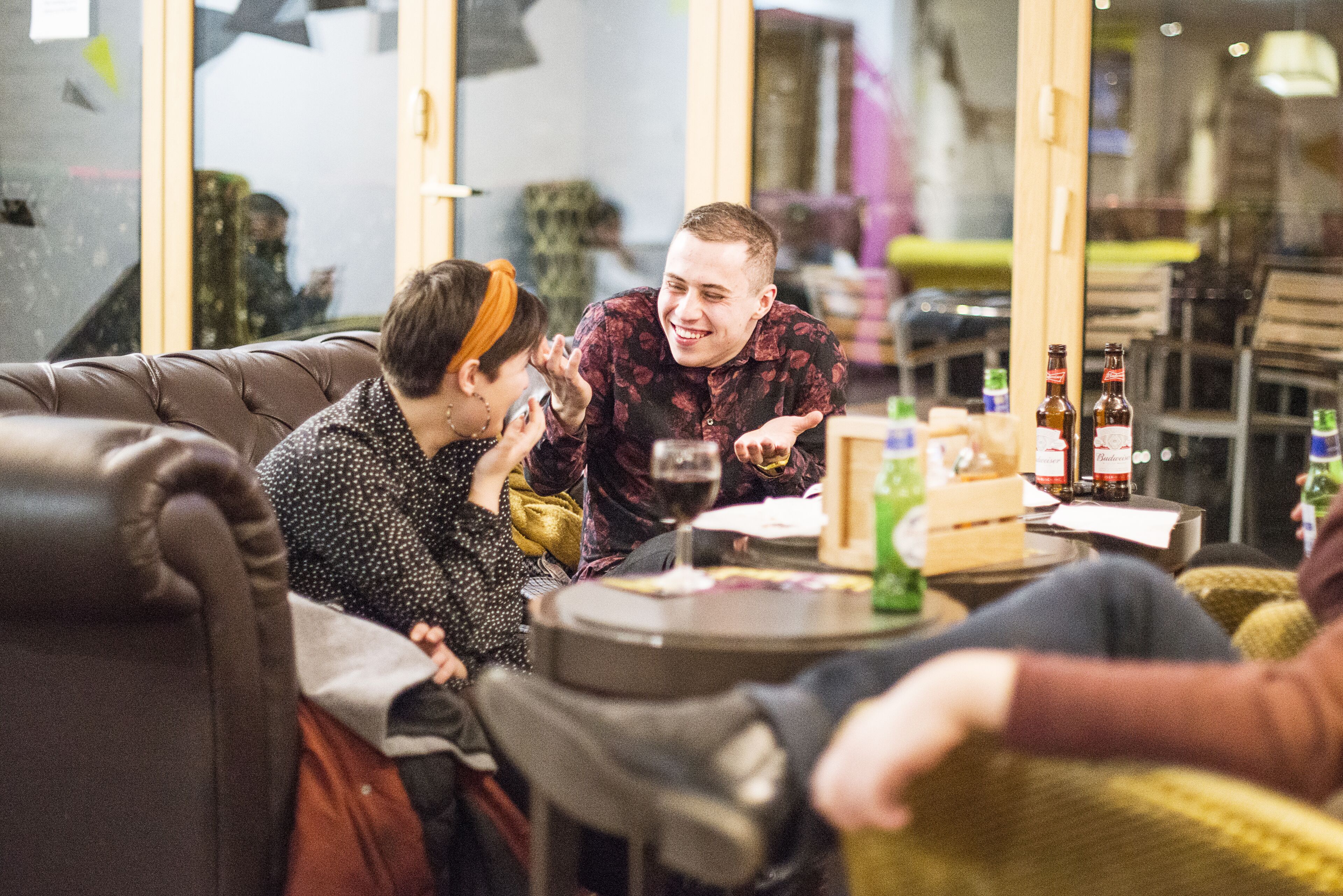 Um grupo de pessoas sentadas num sofá num espaço moderno com paredes de vidro, uma mesa redonda com bebidas e uma janela visível.