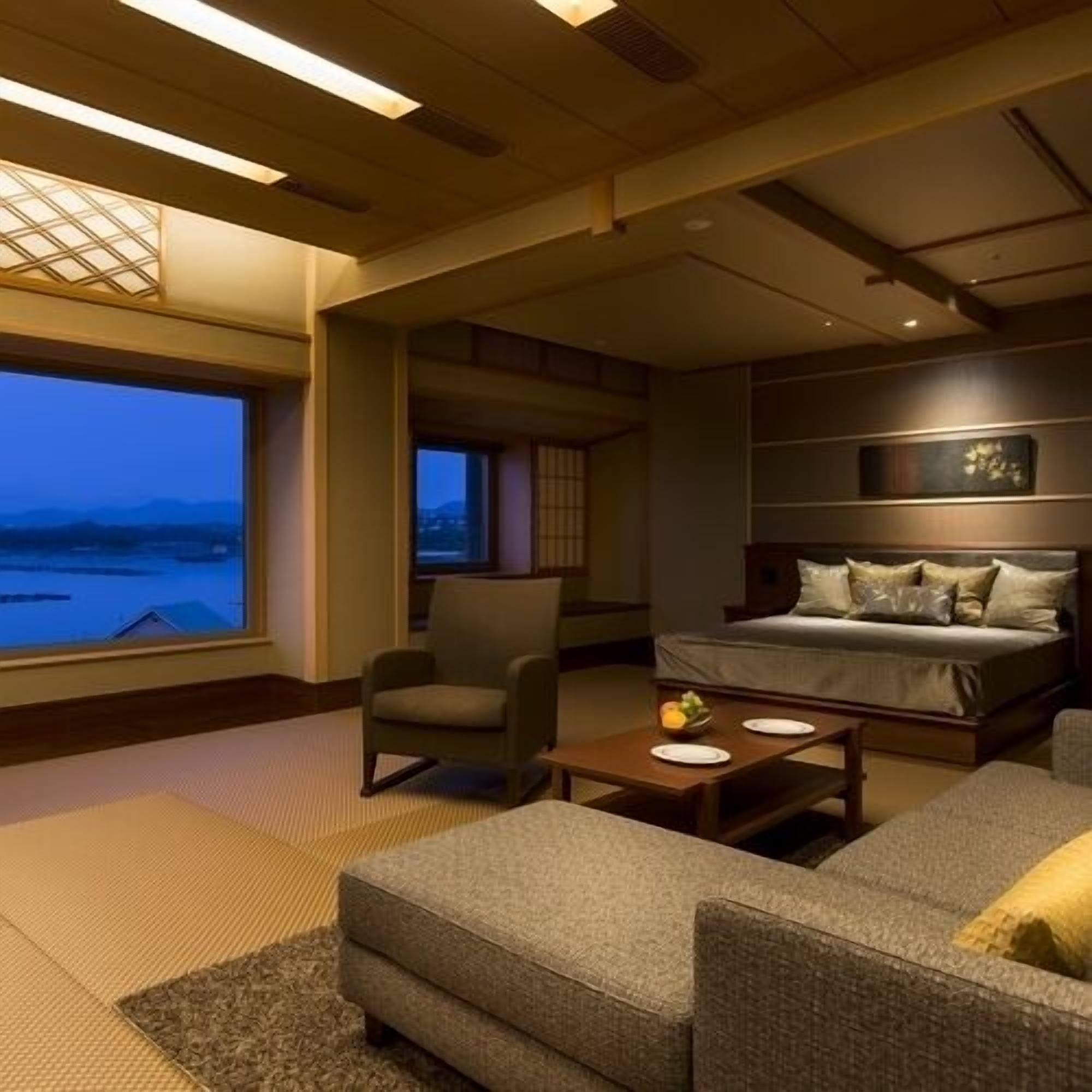 Quarto de hotel moderno com cama, sofá e poltrona, uma mesa de jantar com pratos, e vista para as montanhas.