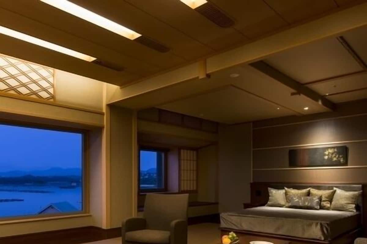 Quarto de hotel moderno com cama, sofá e poltrona, uma mesa de jantar com pratos, e vista para as montanhas.