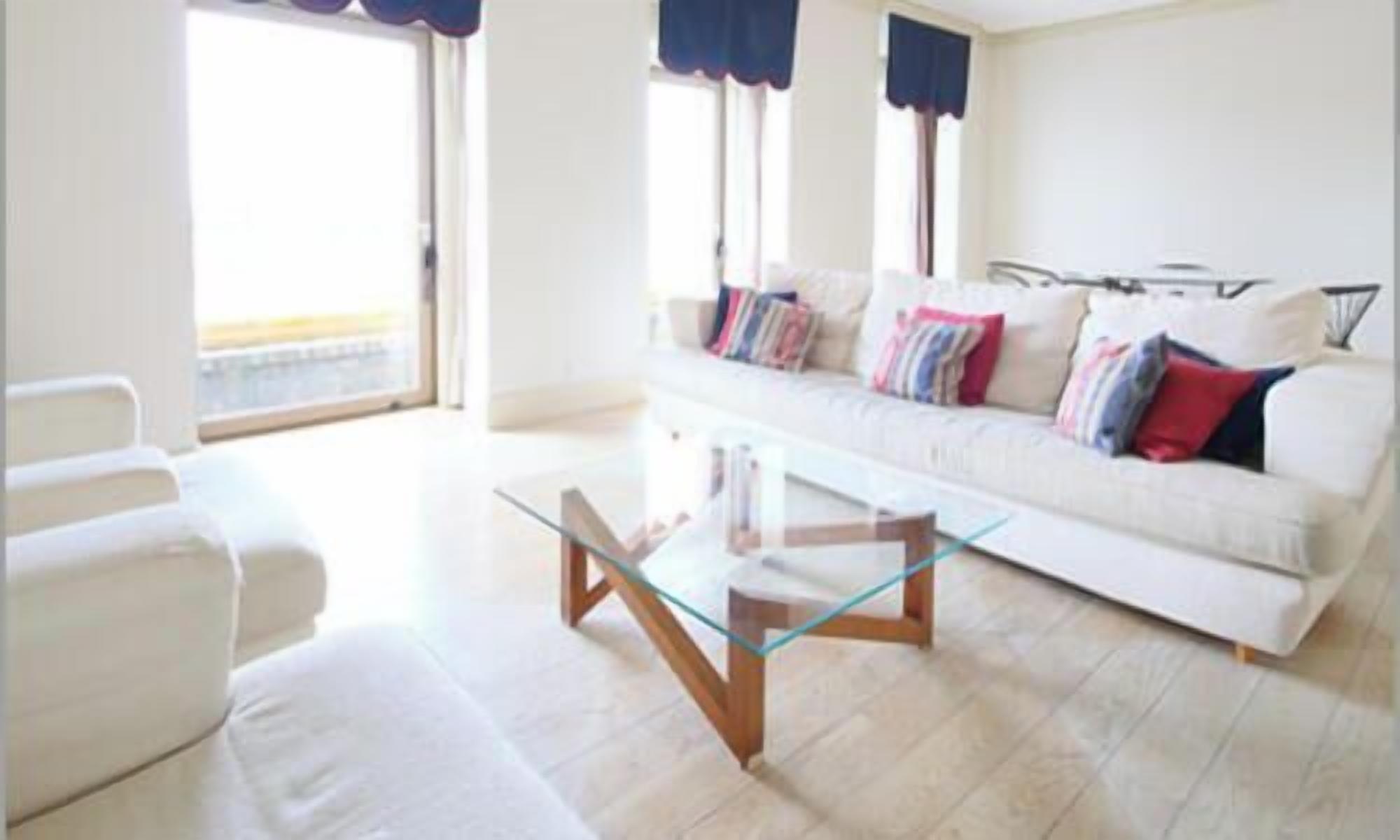 Uma sala de estar com sofá branco, mesa de centro de vidro e vista pela grande janela.