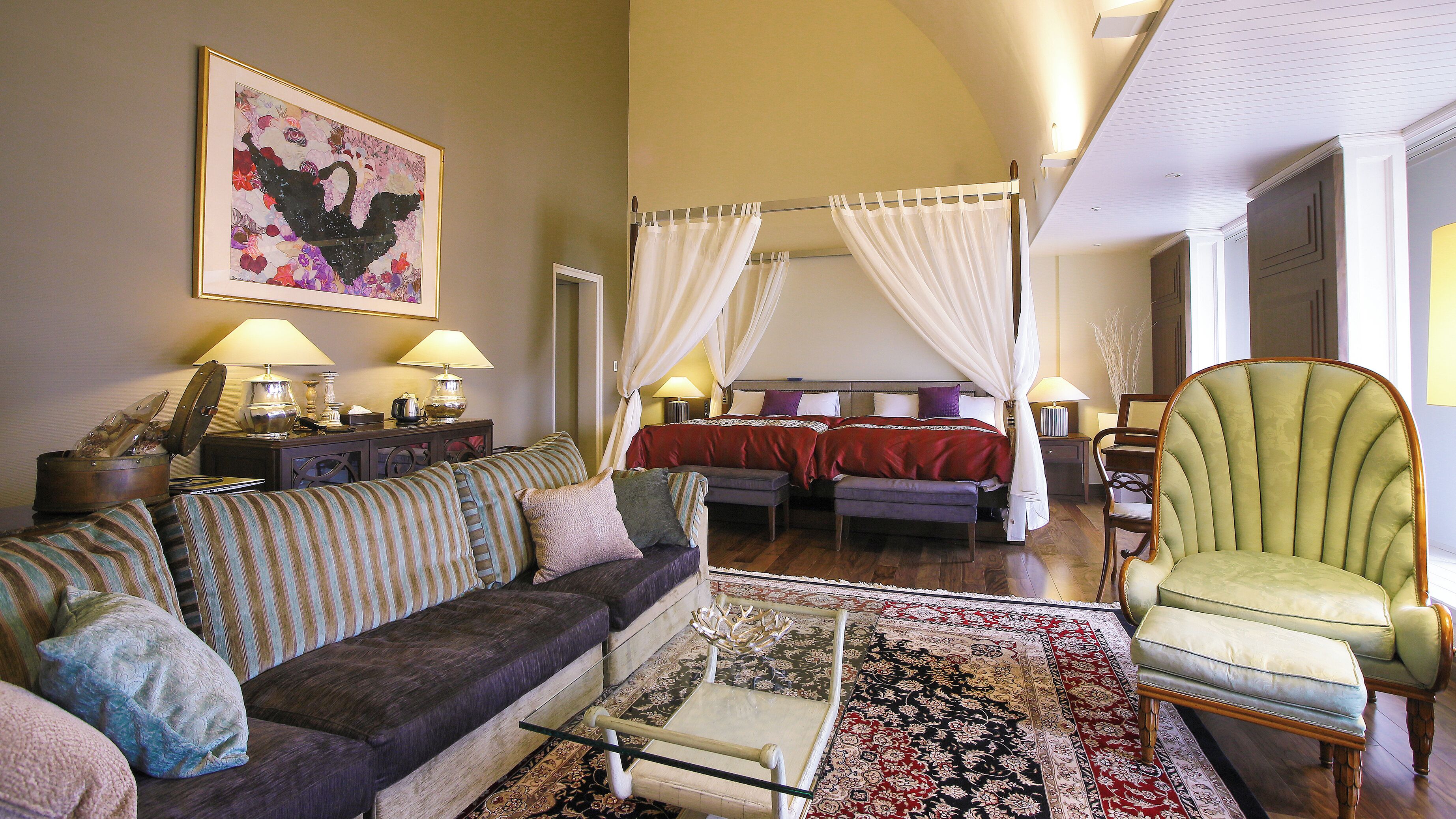 Quarto com cama, área de estar com sofá, poltrona, mesa de cabeceira e mesa lateral com luminárias.