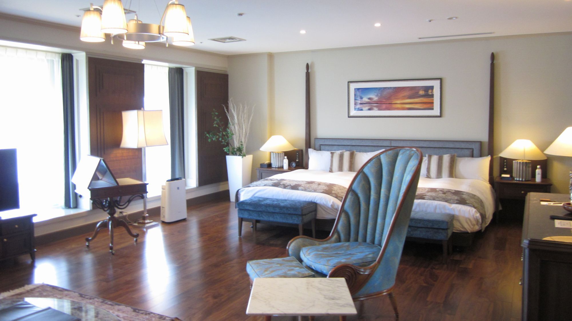 Quarto de hotel com cama, poltrona, escrivaninha, luminária, planta e pintura na parede.