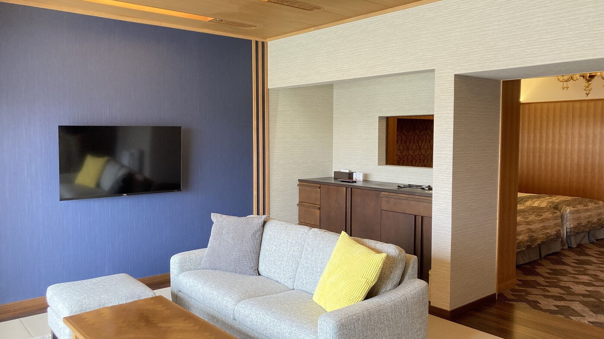 Quarto de hotel com sofá cinzento, mesa de centro de madeira, televisão de tela plana, pequeno espaço de cozinha e carpete estampado.