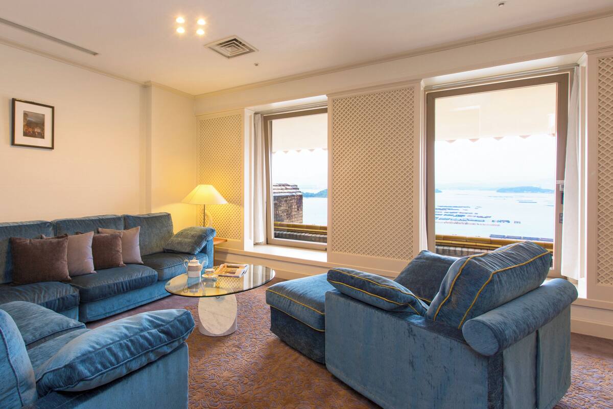 Uma sala de estar com um sofá azul, uma mesa de centro de vidro e vista para o mar através de grandes janelas.
