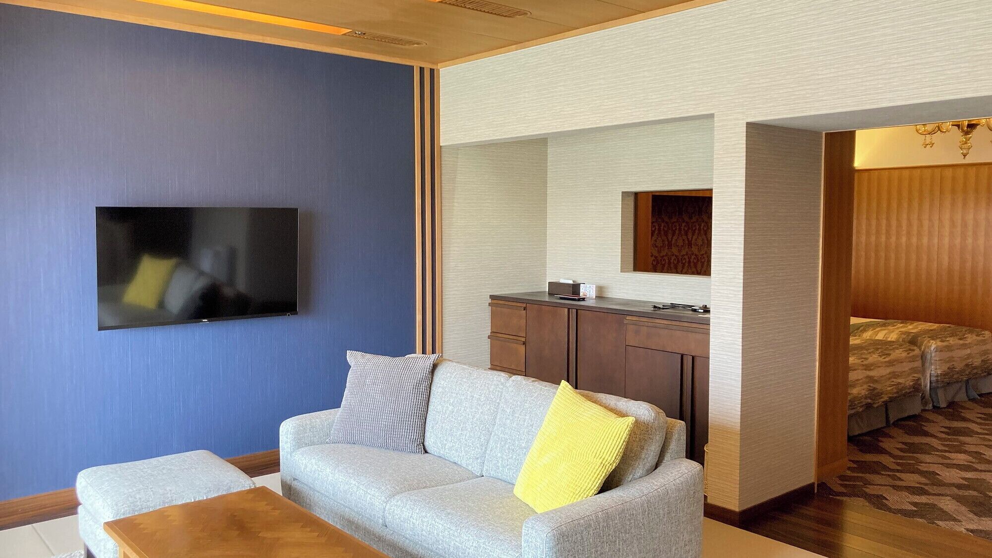 Quarto de hotel com sofá cinza, mesa de centro de madeira, televisão fixada na parede, pequena escrivaninha com espelho e tapete estampado.