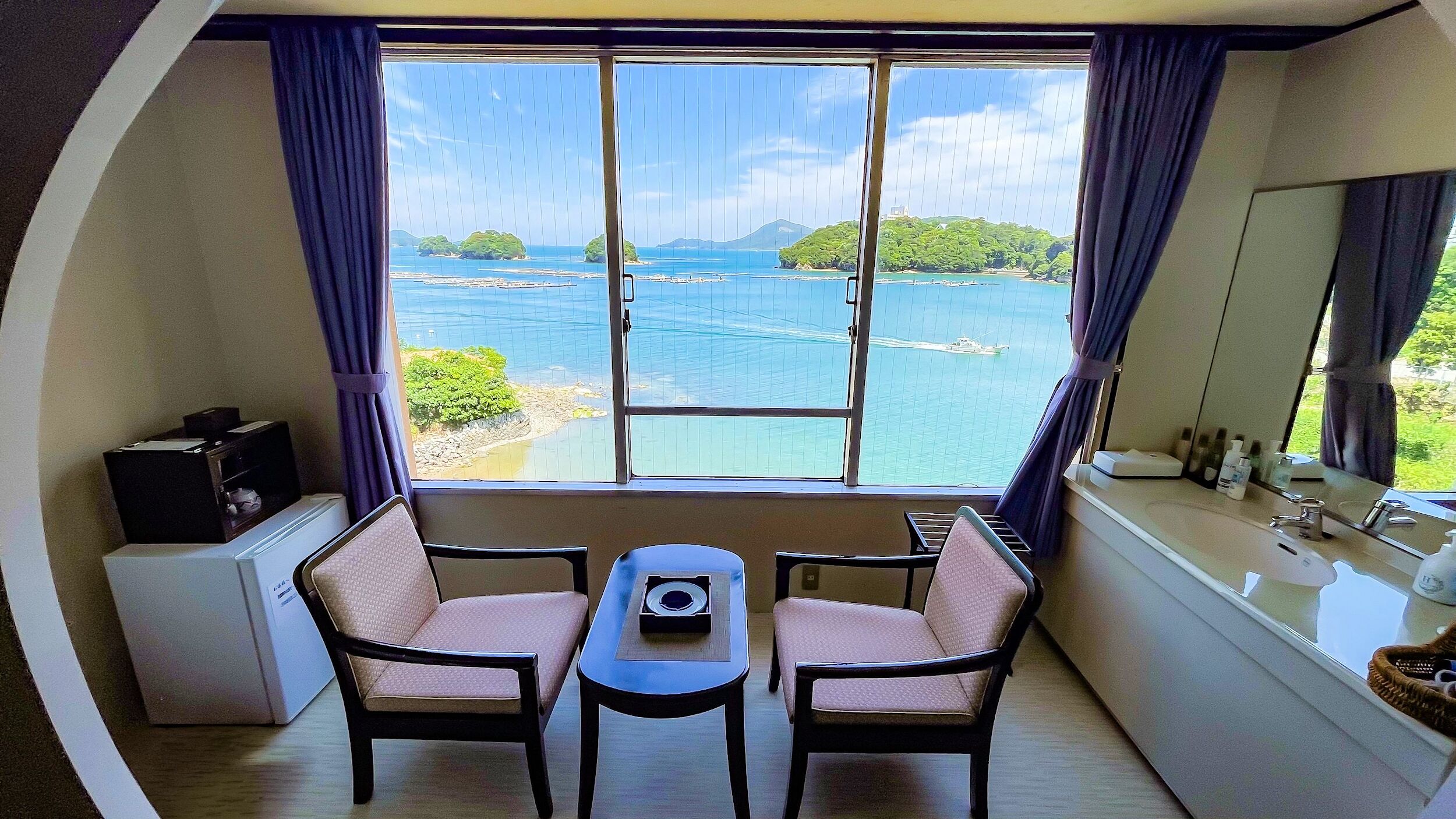 Quarto com uma janela grande que oferece vista para um corpo d'água e vegetação, duas cadeiras, uma mesinha e uma pia com amenities.
