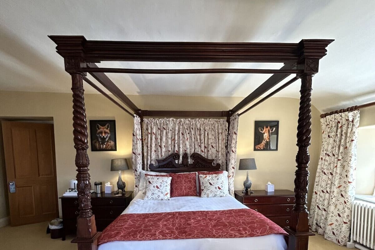 Uma cama com dossel em um quarto com piso coberto por carpete, uma porta de madeira e duas luminárias fixadas na parede.