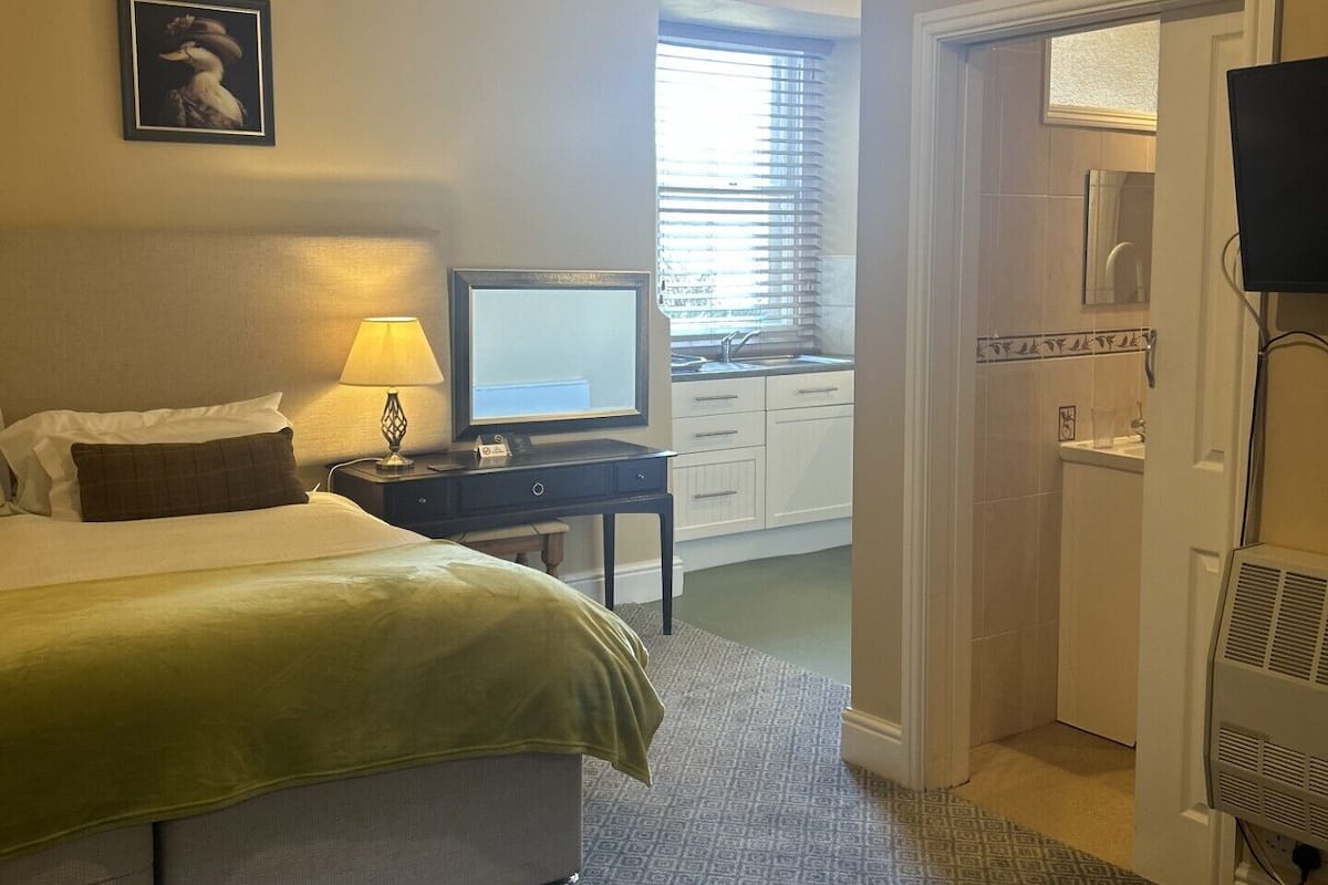 Quarto de hotel com cama, escrivaninha com luminária, televisão e banheiro visível através de uma porta aberta.