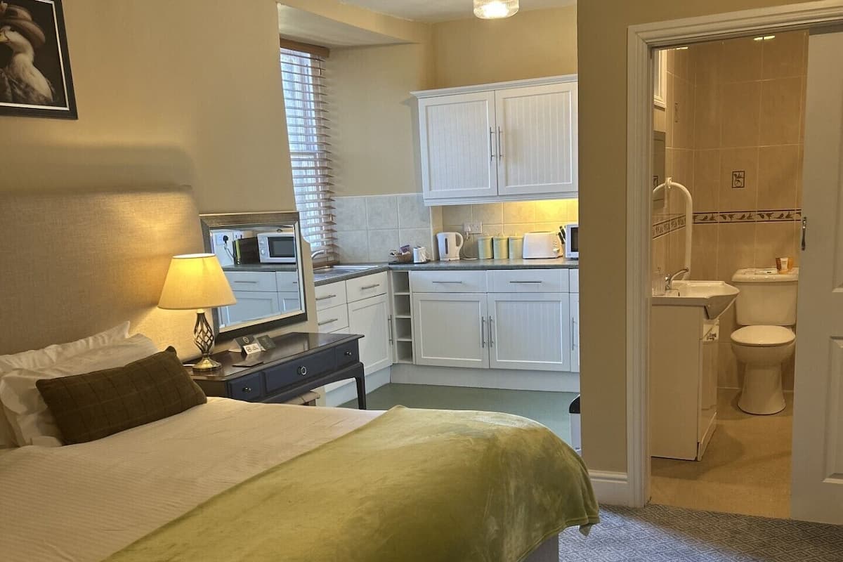 Quarto de hotel com cama, escrivaninha iluminada por uma luminária, cozinha compacta e banheiro.