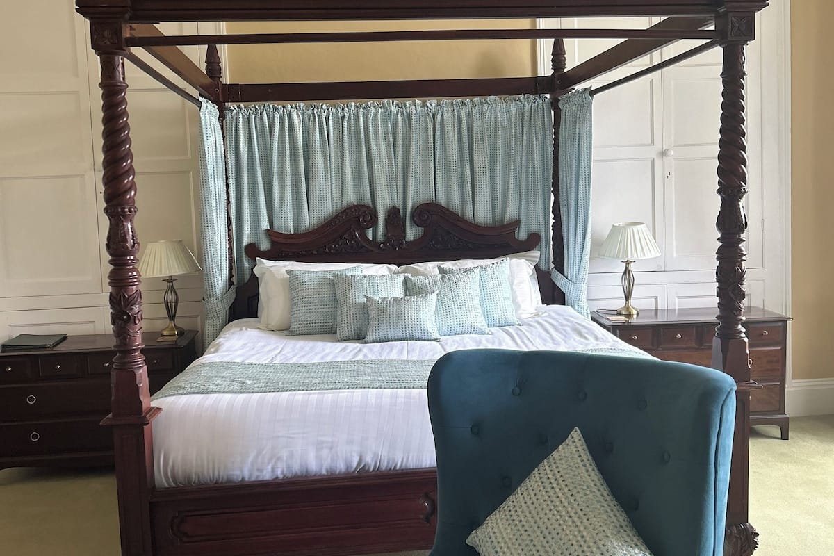 Uma cama com dossel e cortina, uma poltrona estofada azul e uma mesinha de cabeceira com abajur.