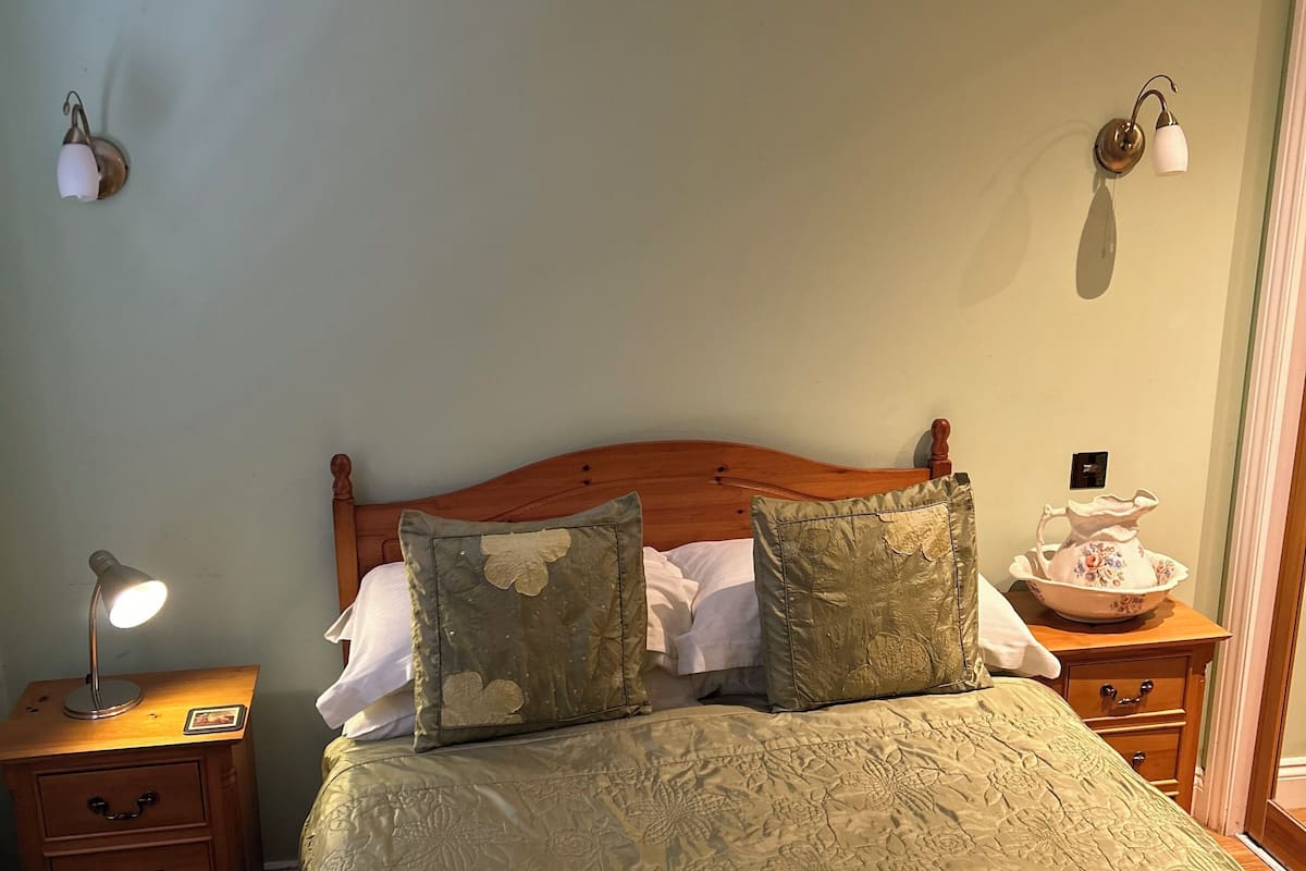 Quarto com cabeceira de madeira, colcha estampada com flores, duas almofadas iguais, mesa de cabeceira com abajur e criado-mudo com uma tigela decorativa.