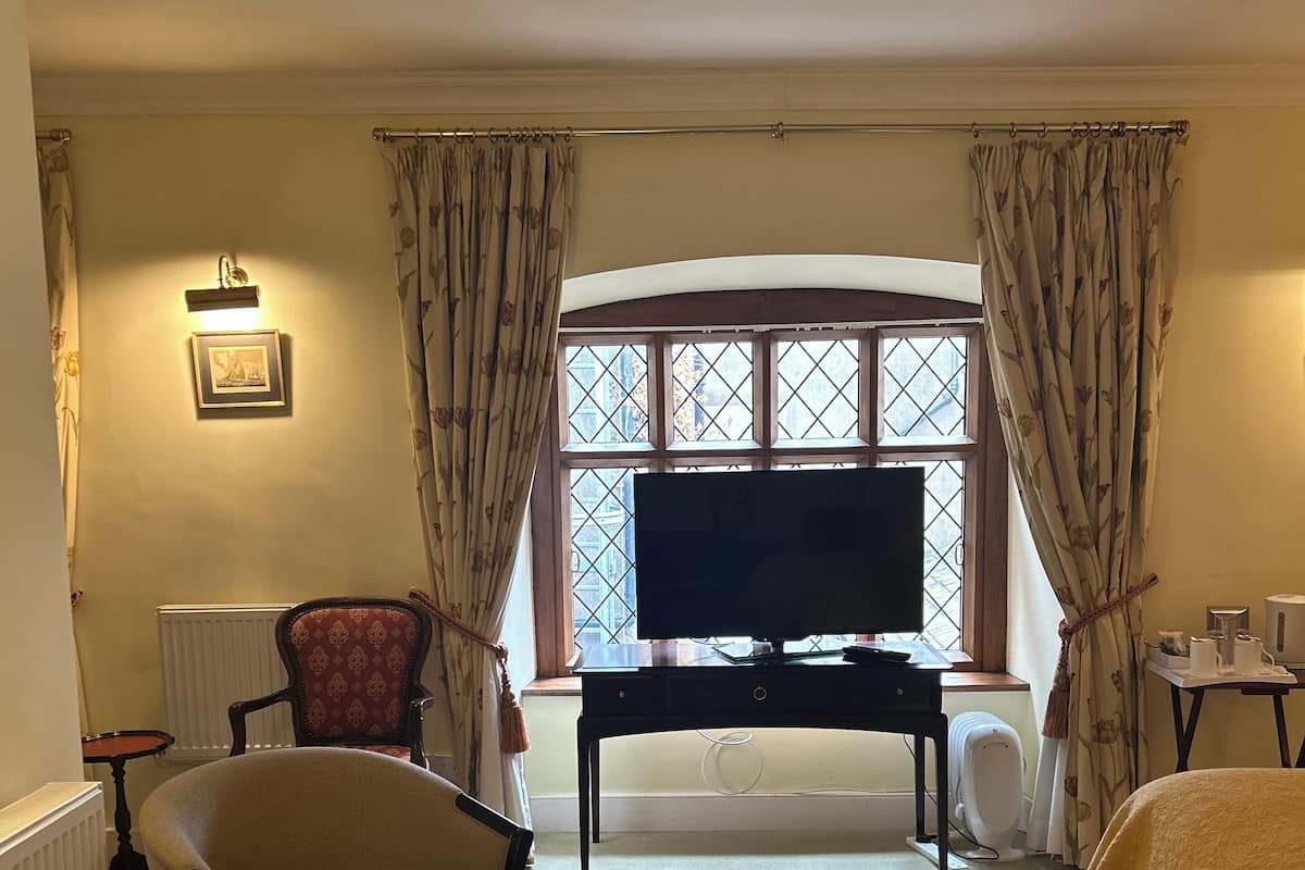 Quarto de hotel com cama, escrivaninha com televisão, cadeira, mesa pequena e radiador.