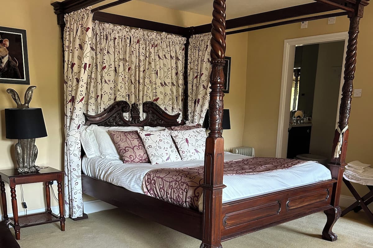 Uma cama de dossel com cortinas florais, uma mesinha de cabeceira com abajur e uma poltrona roxa com almofada floral.