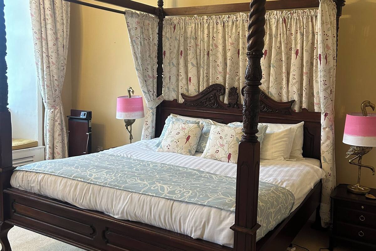 Uma cama com dossel adornada com cortinas florais, uma mesinha de cabeceira com abajur e um criado-mudo com lâmpada.