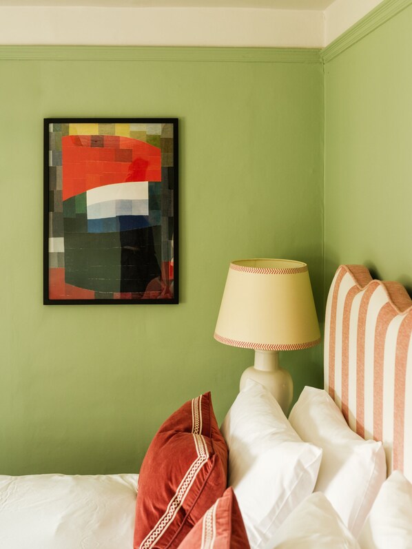 Quarto com parede verde, pintura abstrata emoldurada, luminária de cabeceira e cama com almofadas brancas e listradas.
