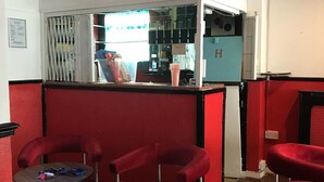 Quarto com cadeiras estofadas em vermelho e uma pequena mesa, área de kitchenette com frigorífico e fogão, e teto com iluminação embutida.