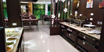 Restaurante com várias mesas e cadeiras, área de buffet com diversos pratos e parede decorativa com motivos florais.