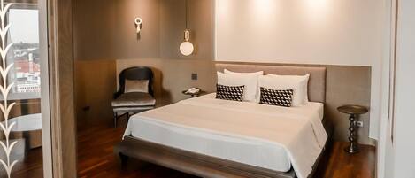 Quarto de hotel com uma cama grande, uma cadeira, mesa de cabeceira e luminária fixada na parede.