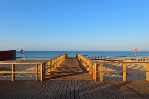 Um calçadão de madeira que leva a uma praia, com aviso de boia salva-vidas e sinalização de proibição de banho.
