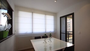 Uma zona de refeições moderna com mesa e cadeiras de vidro, janela com persianas e vista para um edifício.