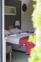 Cama bem arrumada com manta vermelha, uma cadeira de madeira, abajur e espelho na parede.