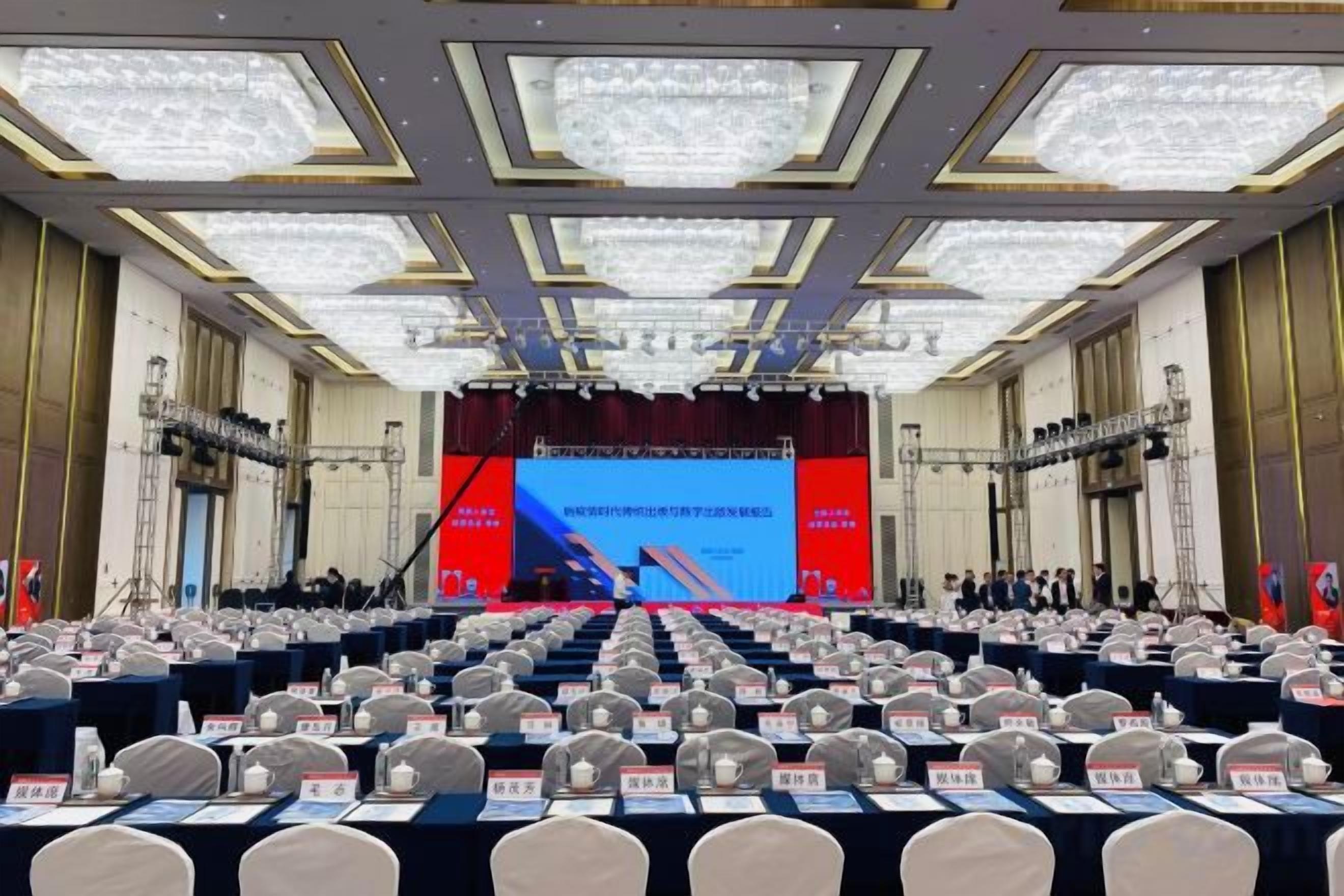 Sala de conferências com mesas alinhadas, palco com uma tela grande e ambiente formal.