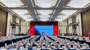 Sala de conferências com mesas alinhadas, palco com uma tela grande e ambiente formal.