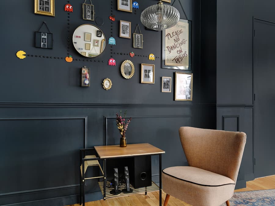 Um quarto com decoração escura, um poltrona de estilo antigo, uma pequena mesa de madeira com um vaso, e uma parede decorada com fotografias emolduradas e objetos decorativos.