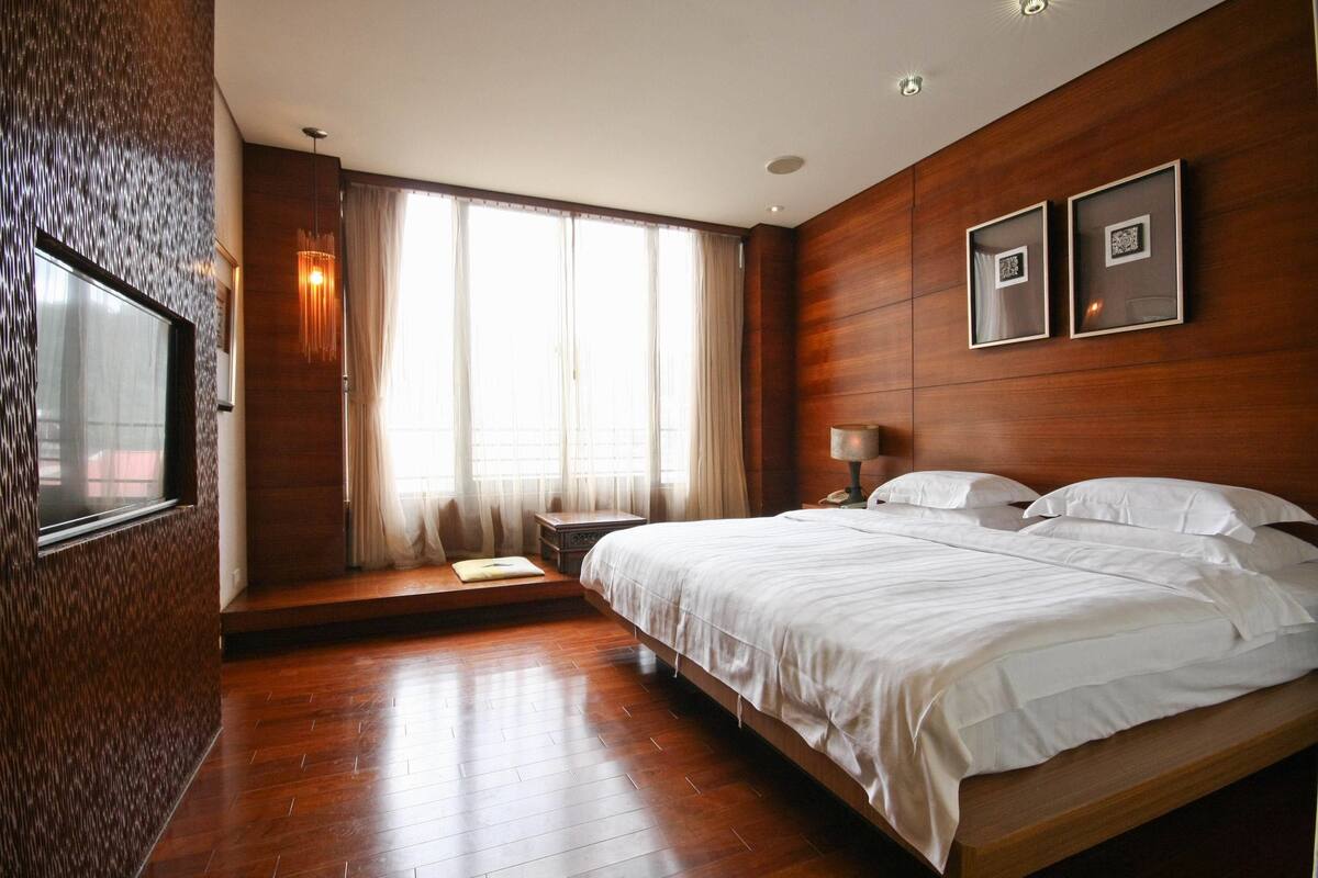 Um quarto com uma cama grande, paredes de madeira, uma janela com cortinas e uma mesa de cabeceira com abajur.