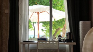 Quarto de hotel com cama, uma mesinha com controle remoto, cafeteira e vista para a praia através de uma janela com cortinas.