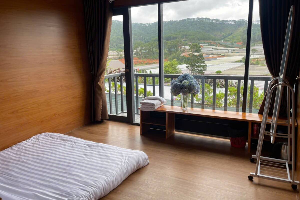 Quarto de hotel com uma cama, uma mesinha com um vaso de flores e vista para uma paisagem montanhosa através de grandes janelas.