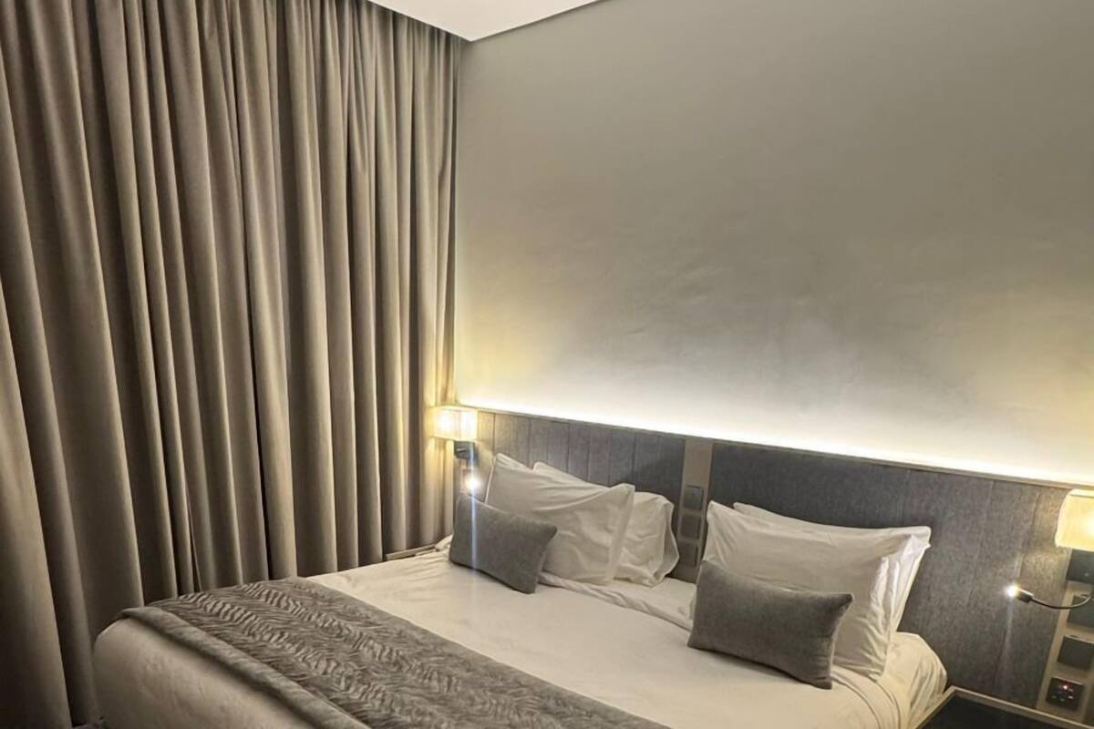 Quarto de hotel com uma cama grande, cabeceira, duas mesas de cabeceira com luminárias e uma cortina na janela.