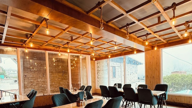 Restaurante com teto alto, vigas de madeira aparentes e lâmpadas penduradas. Mesas e cadeiras dispostas para refeições.