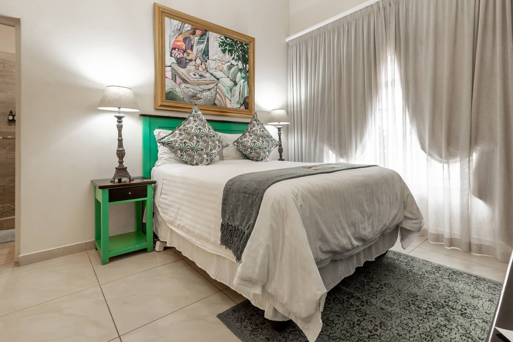 Quarto com cama, duas luminárias de cabeceira, mesa de cabeceira verde, pintura acima da cama e janela com cortinas.