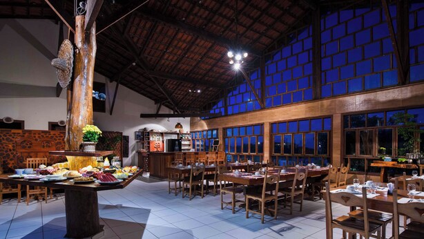 Uma ampla sala de jantar com várias mesas e cadeiras de madeira, um pilar central com decorações penduradas e grandes janelas que oferecem vista do céu noturno.