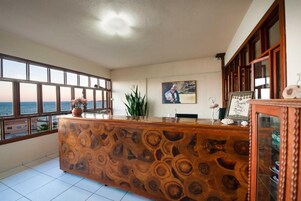 Uma área de recepção com balcão de madeira, janelas amplas com vista para o mar e um quadro na parede.
