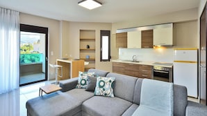 Uma sala de estar moderna com um sofá modular cinza, uma pequena mesa de centro de madeira e almofadas decorativas. Cozinha com fogão, pia e geladeira visível ao fundo.
