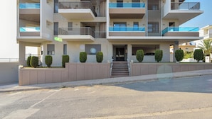 Edifício residencial de vários andares com varandas, entrada central e área de estacionamento em frente.