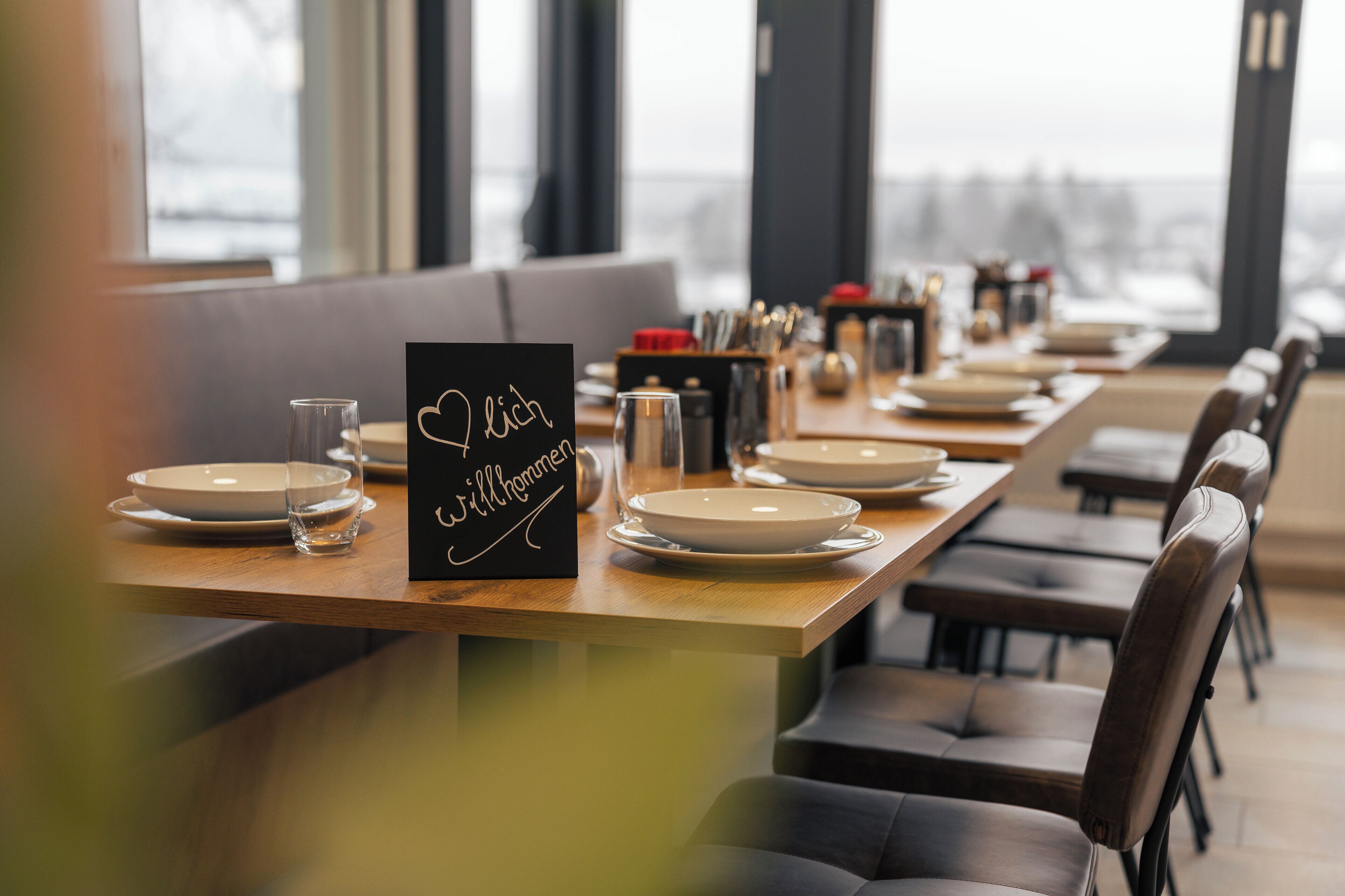 Um ambiente de restaurante com uma mesa comprida posta para refeições, uma placa preta com a palavra 'Willkommen' e vista da cidade através de grandes janelas.