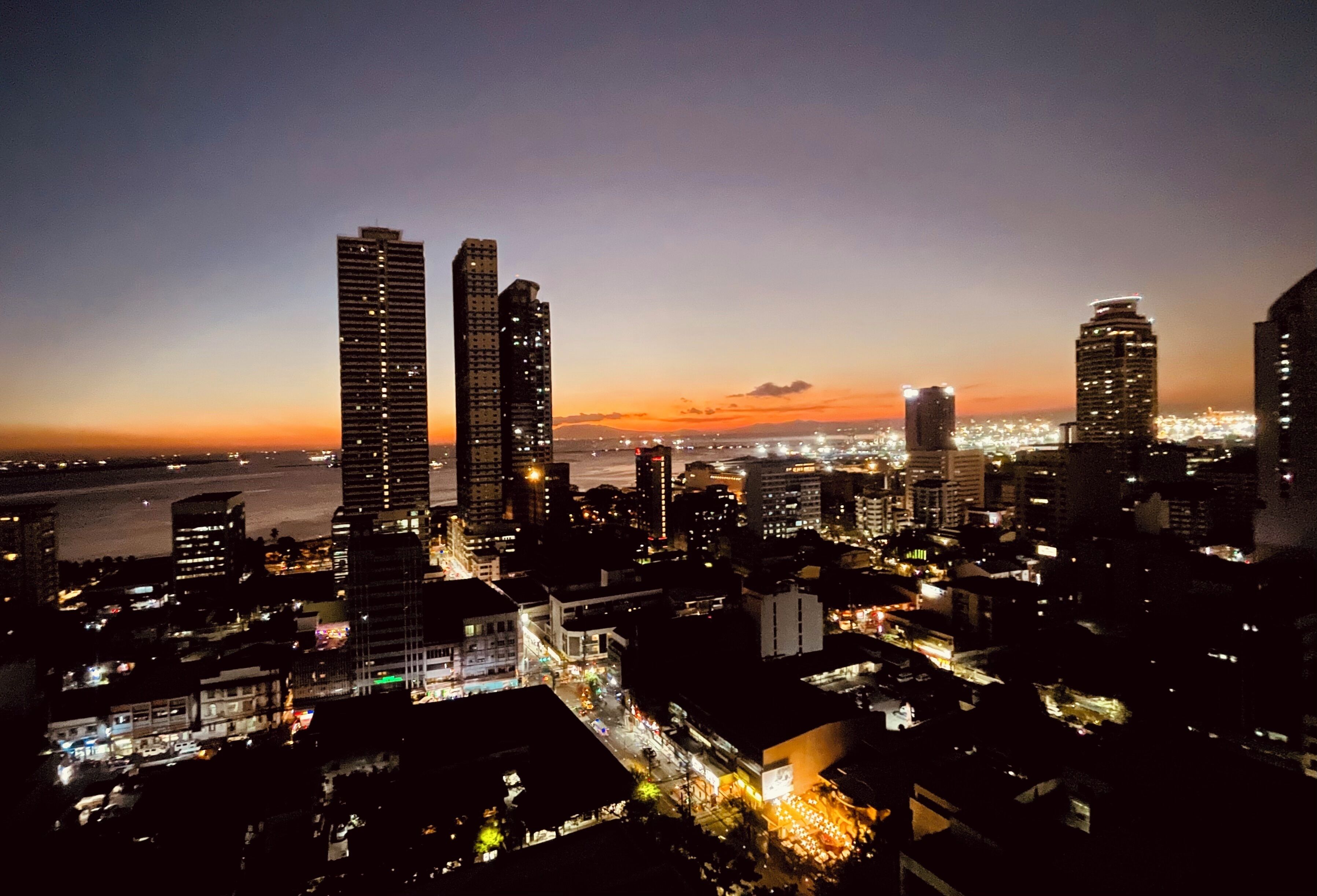 Paisagem urbana ao anoitecer, com edifícios iluminados e um rio ao fundo.
