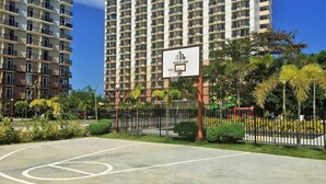Uma quadra de basquete com uma placa, cercada por uma grade e vegetação, com edifícios altos ao fundo.