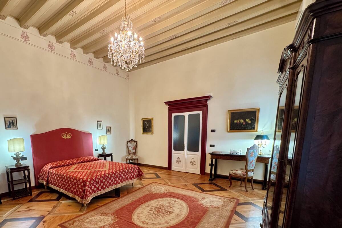Quarto com cama vermelha, piso de madeira, lustre, escrivaninha e uma cadeira.