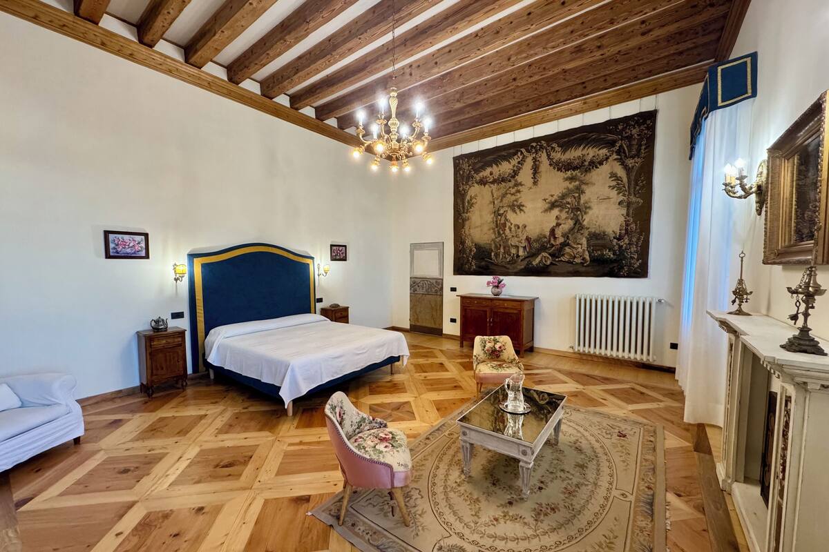 Quarto com uma cama grande, piso de madeira, um lustre, um grande tapeçário e uma lareira clássica.