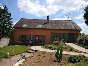 Uma casa de dois andares com telhado de telhas, varanda frontal e um jardim com diversas plantas.