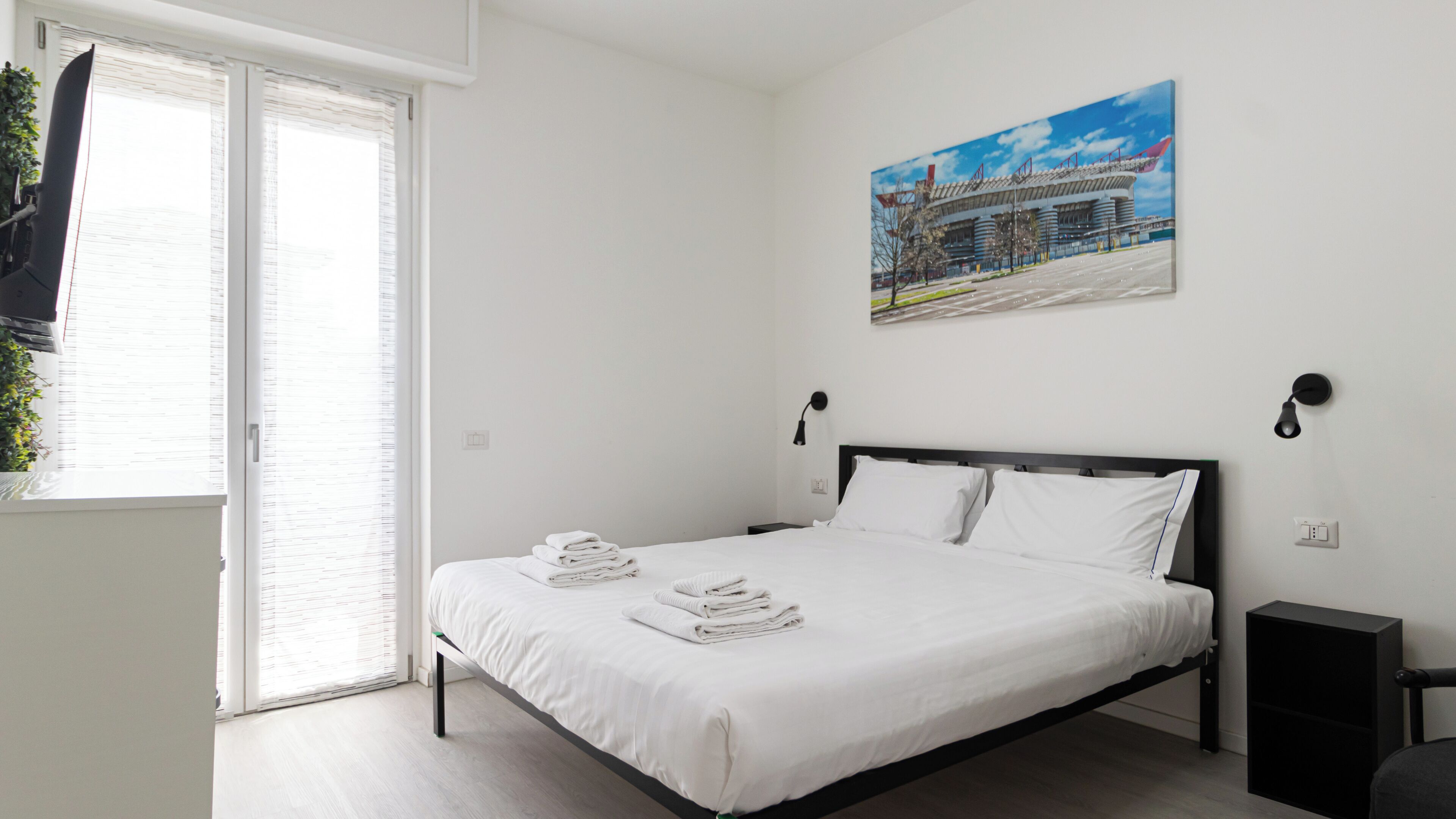 Quarto com cama, pintura de um estádio, janela com persianas e uma planta.