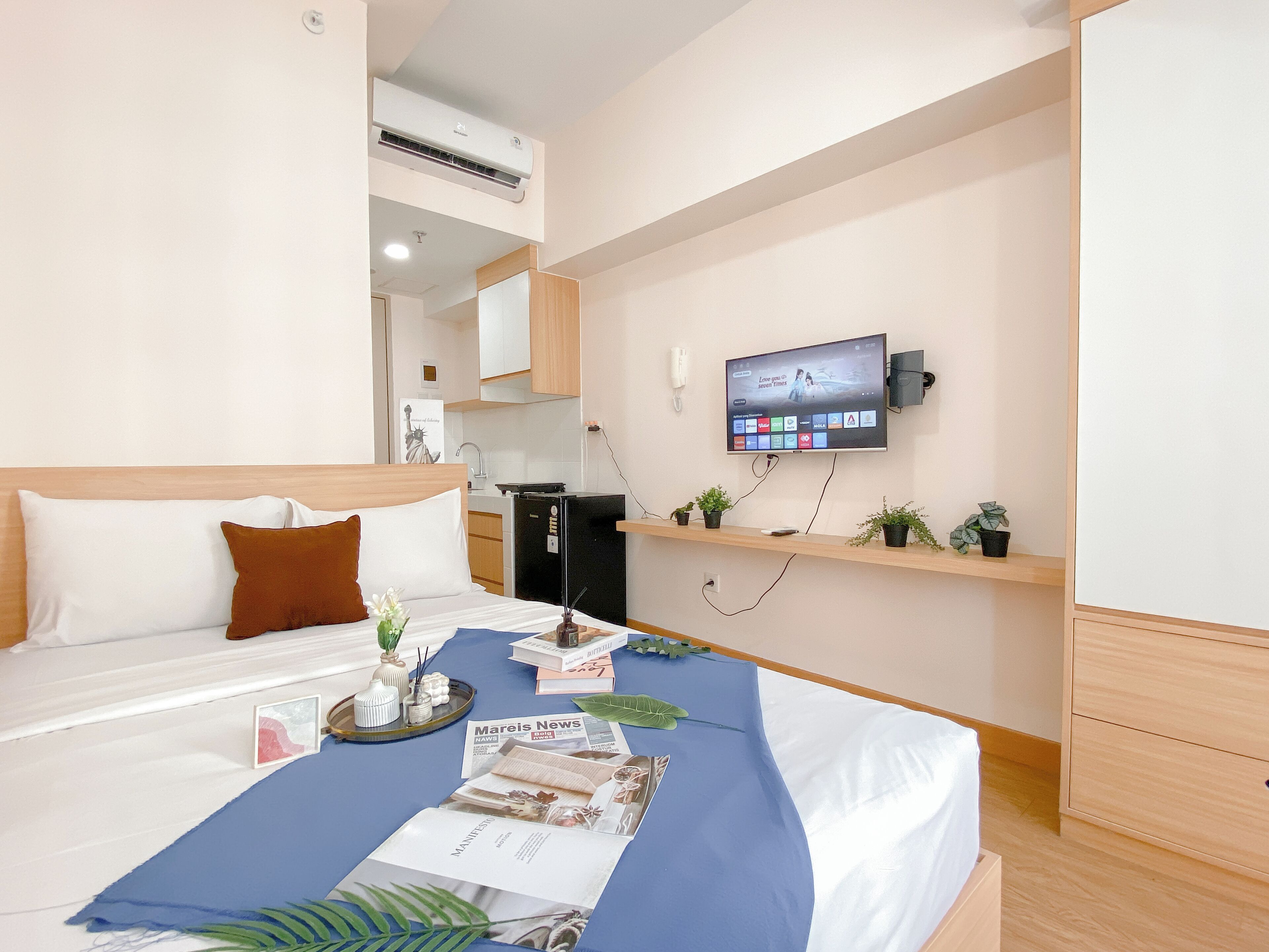 Quarto de hotel com cama, televisão na parede, pequena cozinha e estante com plantas.